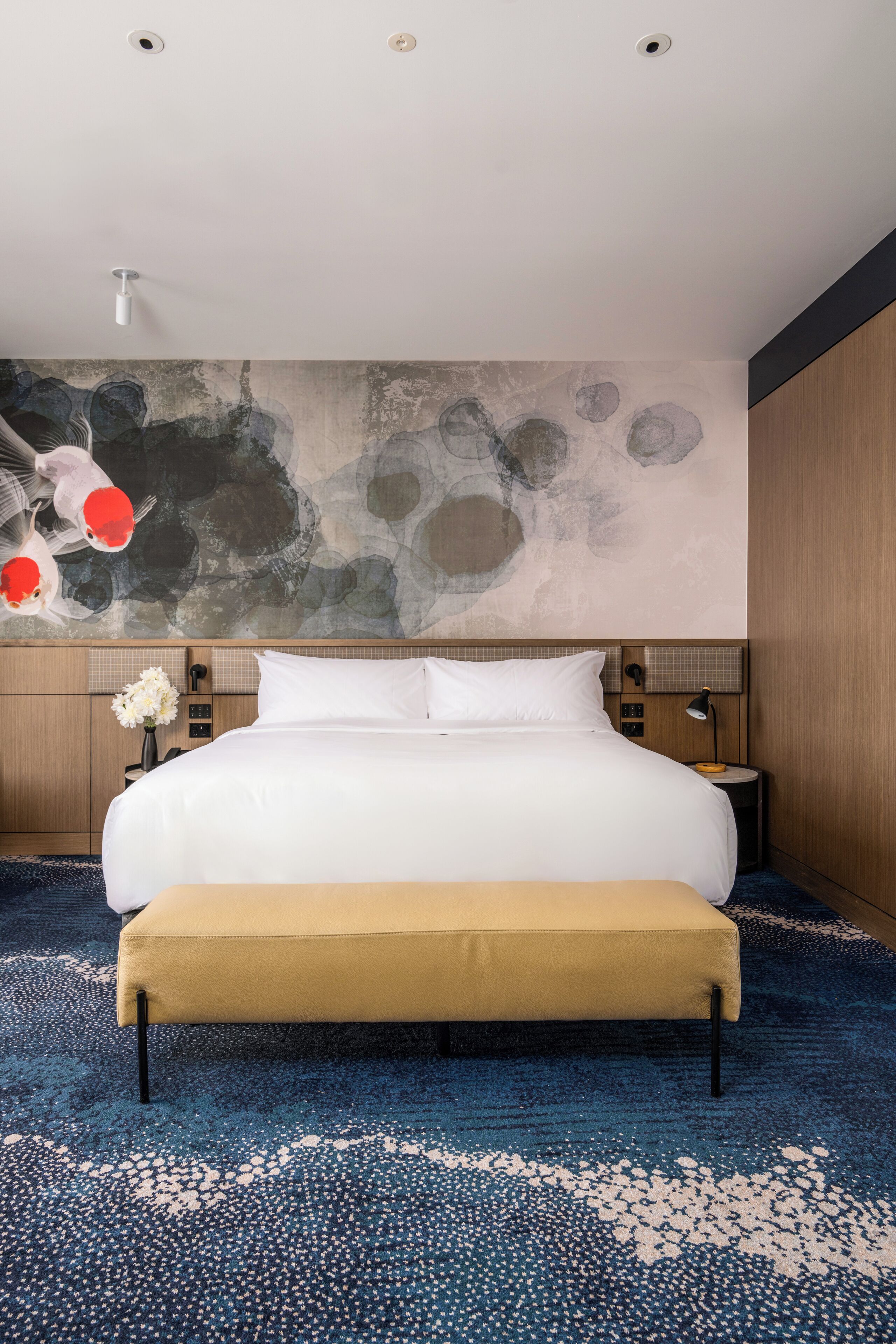 Quarto de hotel moderno com uma cama grande, um banco, uma obra de arte na parede e uma mesa de cabeceira com abajur.