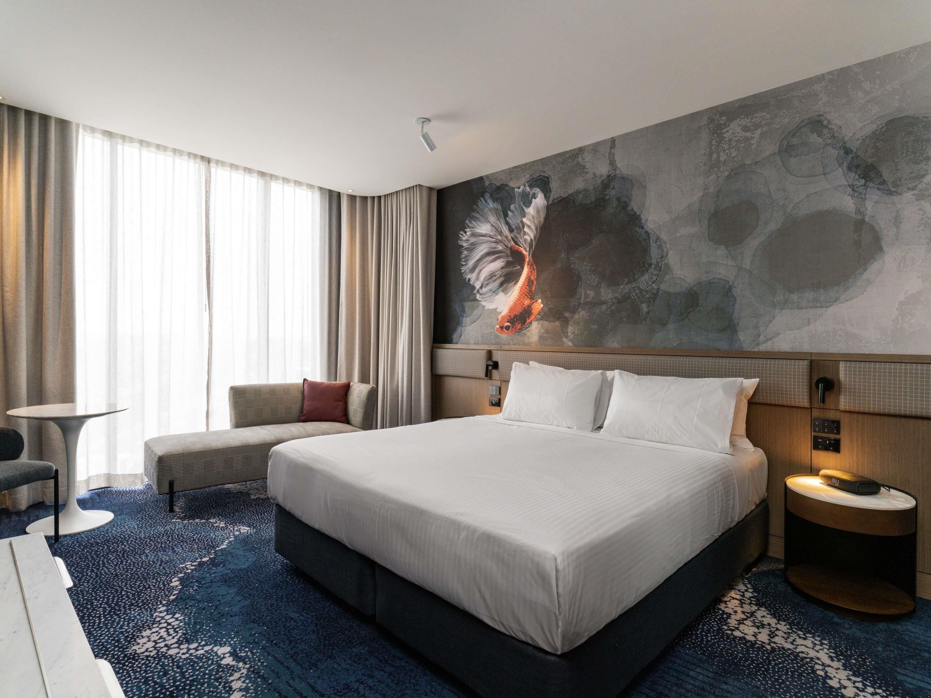 Quarto de hotel com uma cama grande, área de estar com sofá e poltrona, uma mesinha e uma obra de arte na parede.