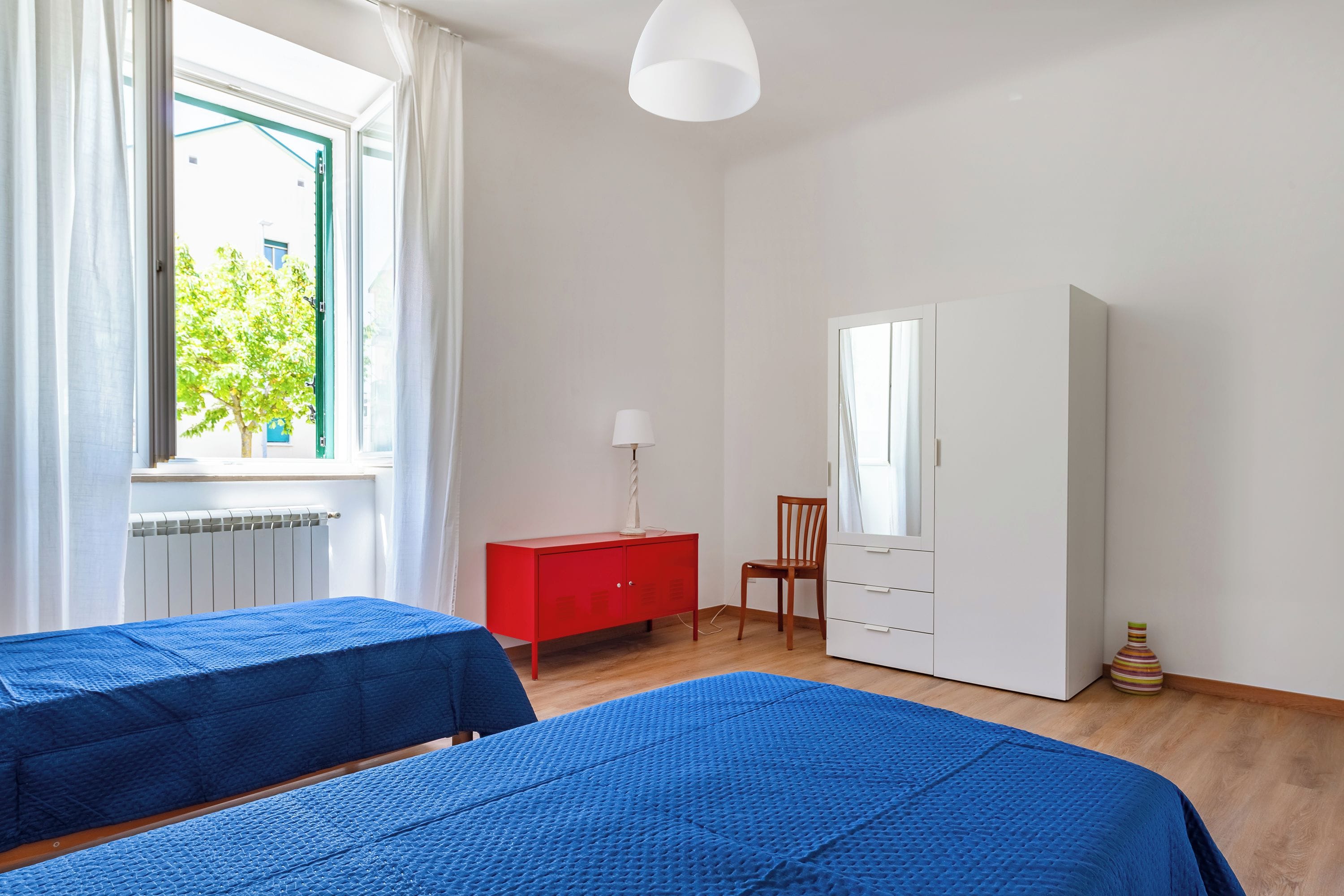 Quarto com duas camas, penteadeira vermelha, guarda-roupa branco e uma janela com cortinas.