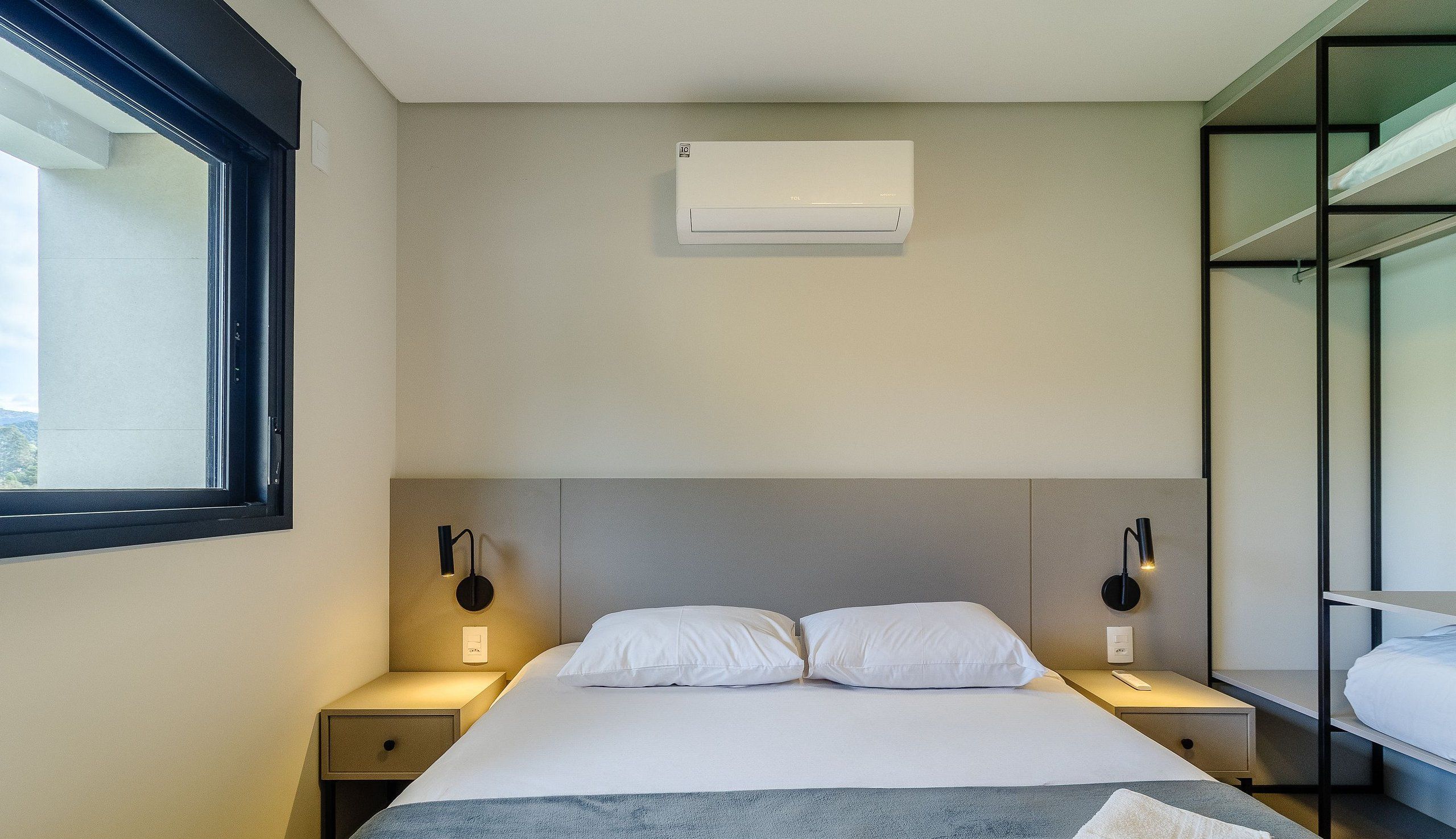 Quarto de hotel moderno com uma cama grande, mesas de cabeceira, ar condicionado fixado na parede e uma janela com vista.