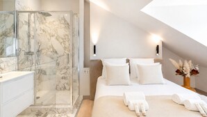 Quarto de hotel moderno com chuveiro em gabinete de vidro, uma cama grande com roupa de cama branca e um vaso com flores secas na mesa de cabeceira.
