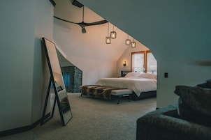 Quarto com teto inclinado, cama com colcha branca, parede de destaque em pedra e luminárias pendentes.