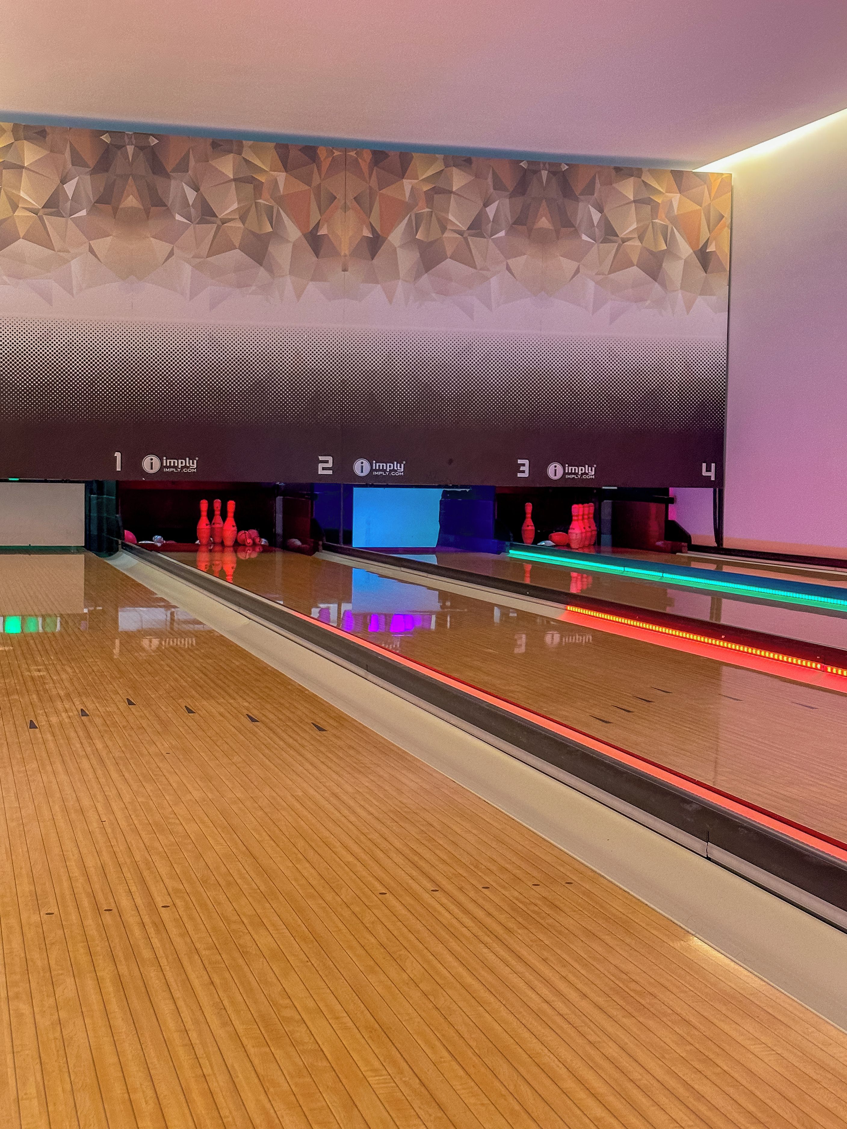 Uma pista de boliche com várias pistas, piso de madeira e uma parede moderna com padrões geométricos.