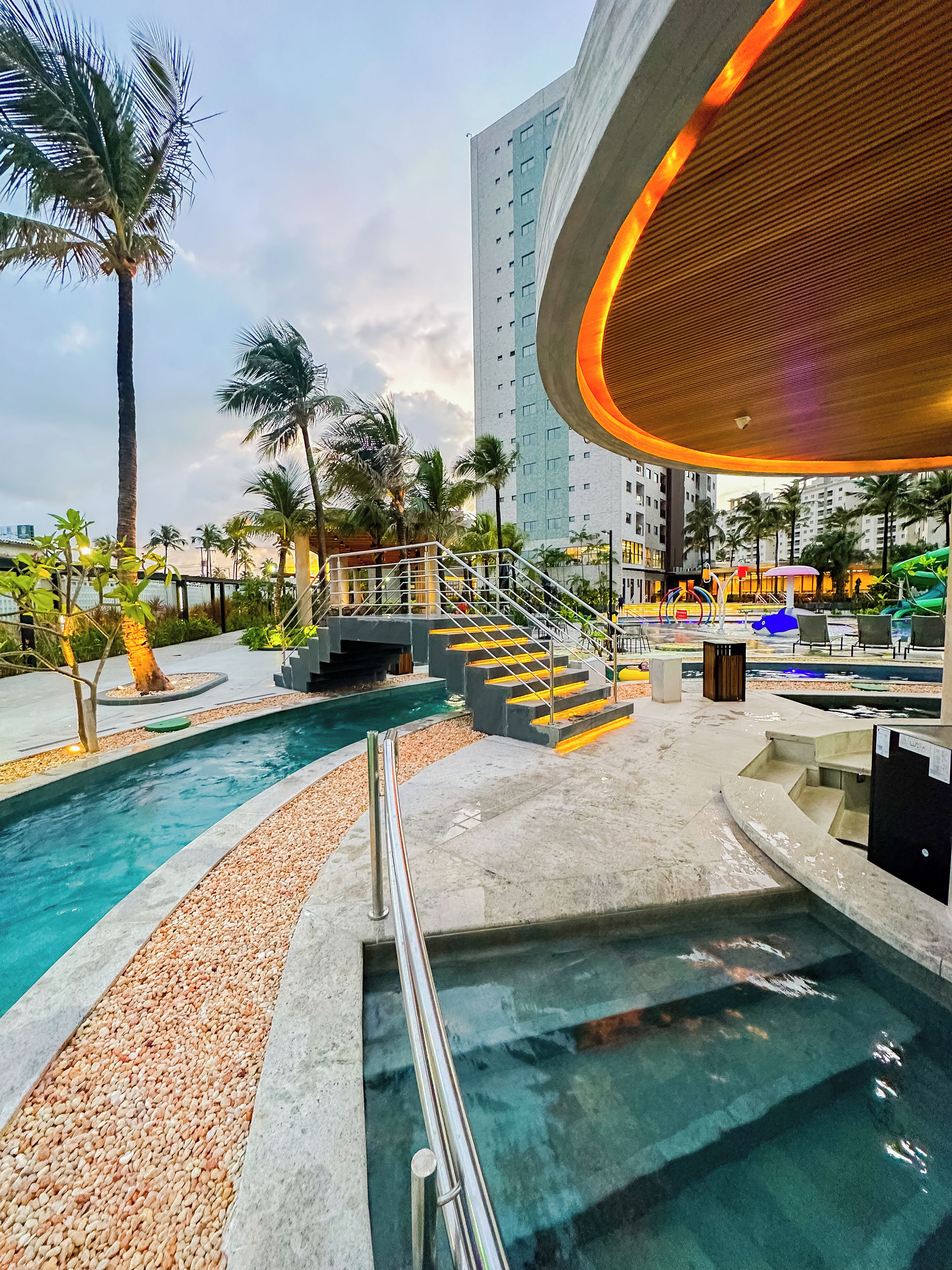 Área de piscina com água cristalina, palmeiras e um edifício moderno ao fundo.