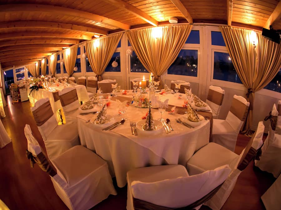Uma sala de banquetes com mesas redondas dispostas para refeições, toalhas brancas e cadeiras com capas brancas.
