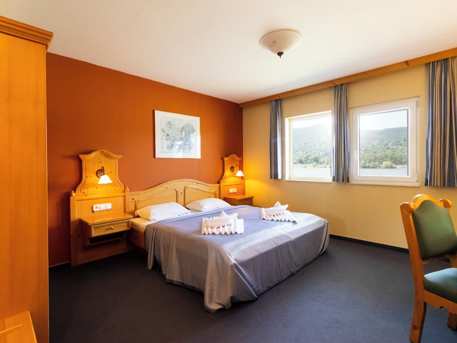 Quarto de hotel com cama, duas mesas de cabeceira com luminárias, uma cadeira e janela com cortinas.