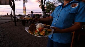 Pessoa segurando uma bandeja com comida, incluindo arroz, feijão e legumes, em um restaurante à beira-mar.