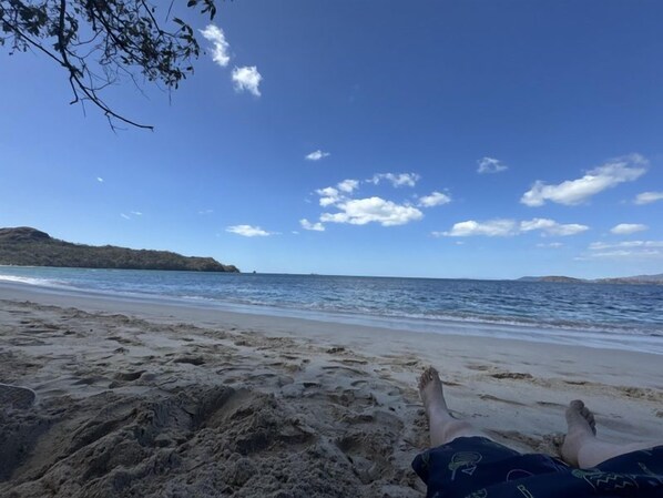 Uma praia com céu claro, areal arenoso e uma pessoa deitada na areia.