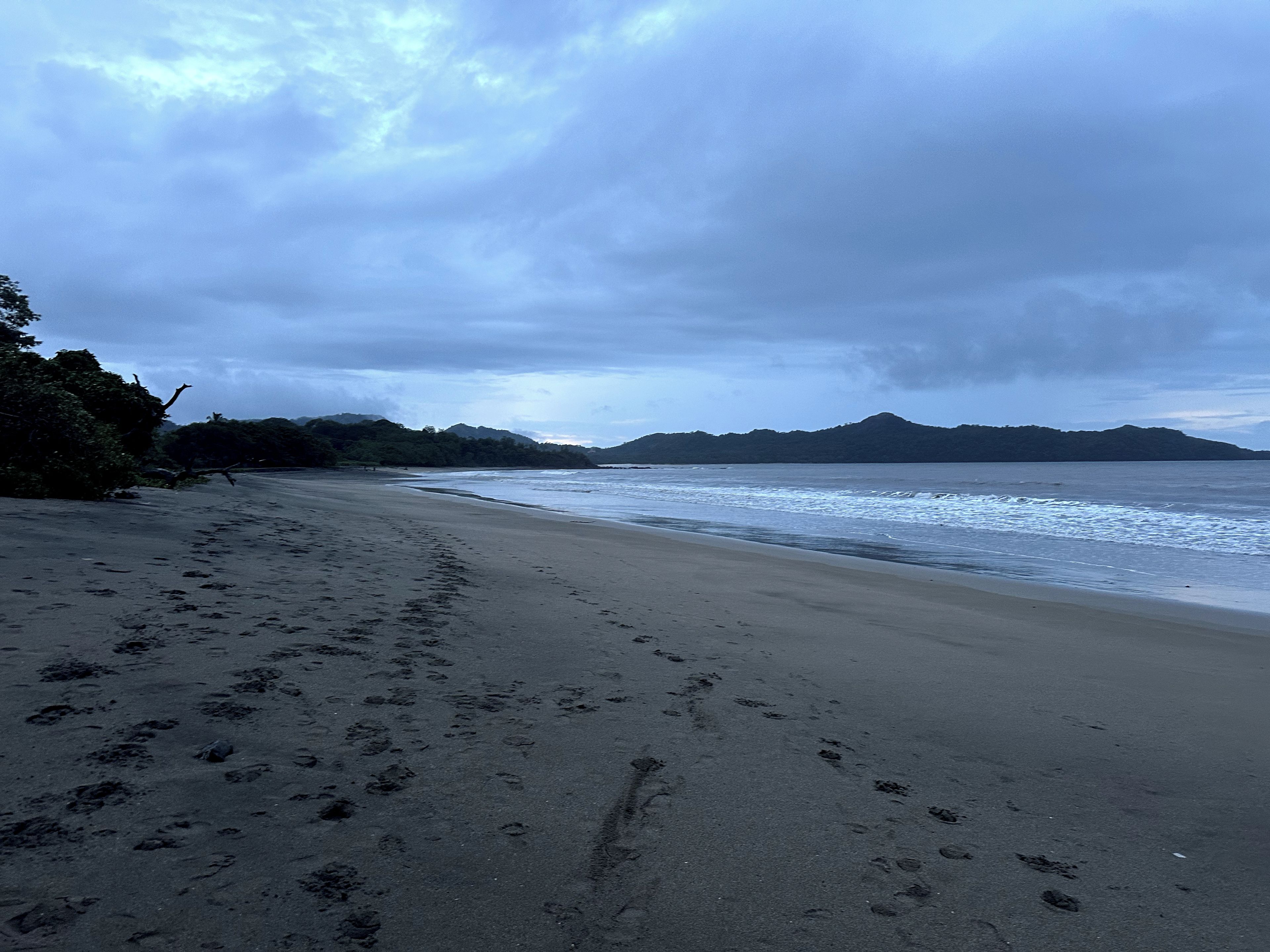 Uma praia com pegadas a levar à água, uma montanha ao longe e céu nublado.