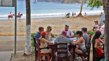 Um grupo de pessoas sentado à mesa de madeira ao ar livre, com uma praia e oceano ao fundo.