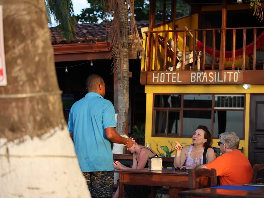 Um hotel chamado 'HOTEL BRASILITO' com uma placa, uma palmeira e uma mesa de madeira com pessoas sentadas.