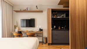 Quarto de hotel com cama, secretária com televisão, cozinha e mesa de jantar.