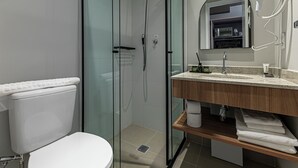 Um banheiro moderno com chuveiro em box de vidro, vaso sanitário branco, toucador com espelho e secador de cabelo fixado na parede.
