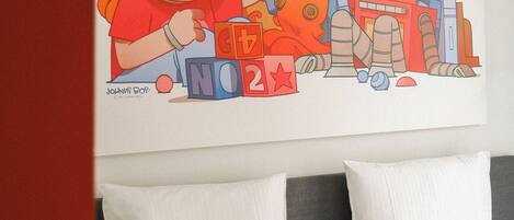 Quarto de hotel com cama, almofadas brancas e obra de arte colorida mostrando uma criança brincando com blocos e robôs.