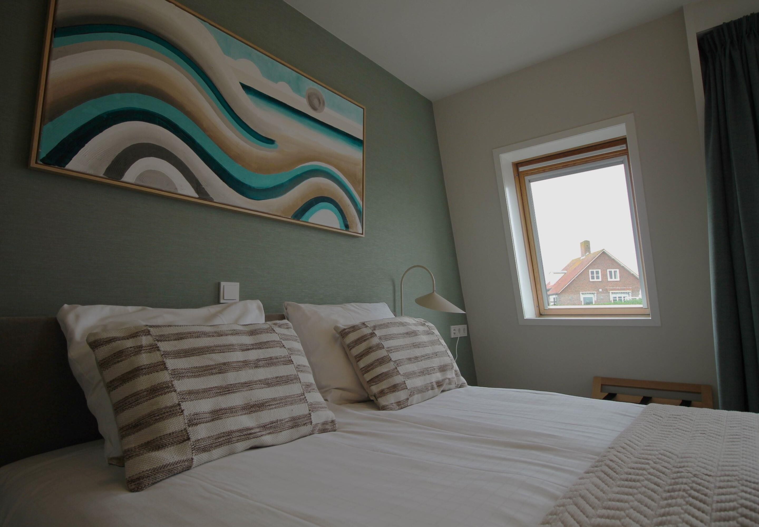 Uma cama bem arrumada com almofadas listradas, um quadro abstrato na parede, uma luminária de cabeceira e uma janela com vista para uma casa.