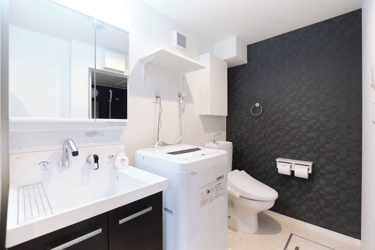 Casa de banho moderna com lavatório branco, espelho e sanita, dispensador de toalhas fixado na parede e parede com textura escura.