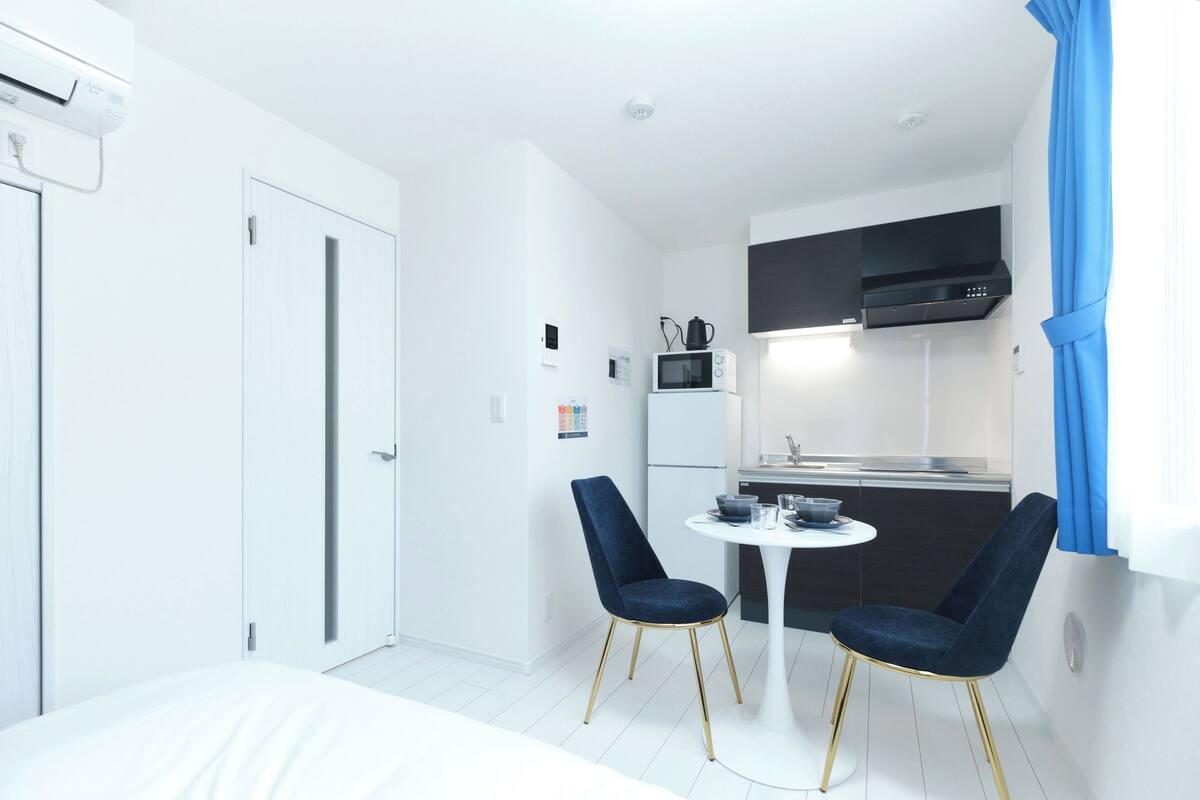 Quarto de hotel moderno com uma cama, duas cadeiras, uma pequena mesa, uma cozinha compacta e uma cortina azul.