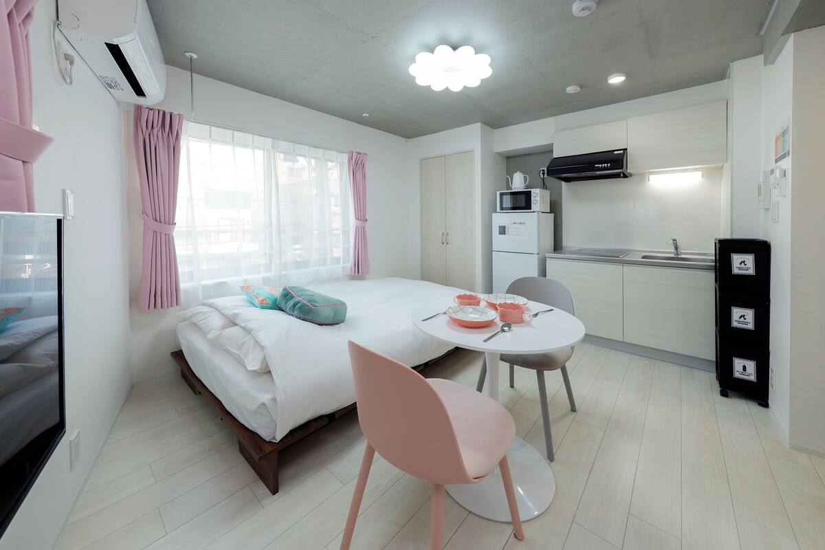 Quarto de hotel moderno com cama, mesa de jantar e cadeiras, cozinha compacta e janela com cortinas.