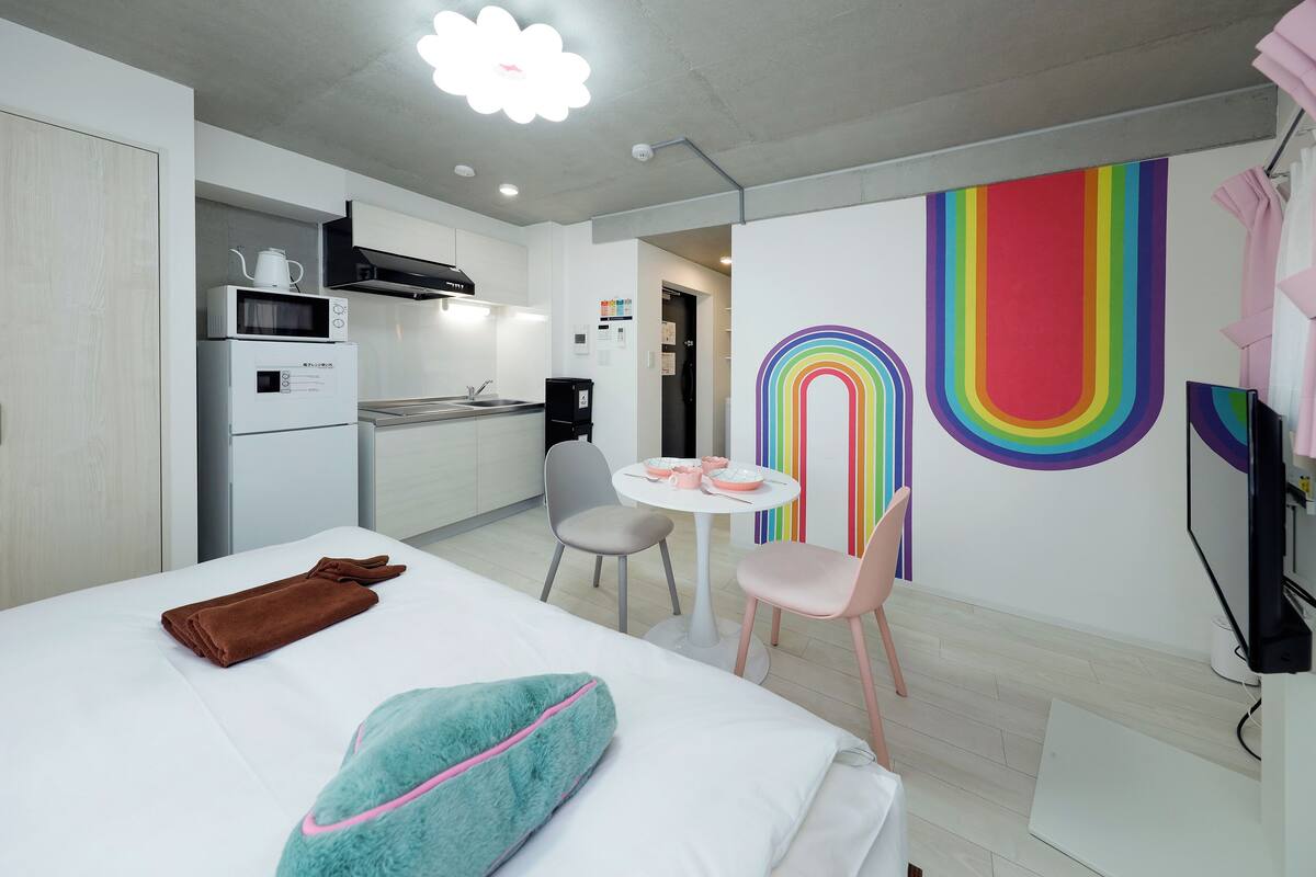 Quarto de hotel moderno com uma cama, uma mesa pequena, uma cadeira e uma cozinha compacta.