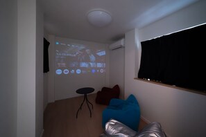 Quarto com tela de projeção exibindo uma apresentação, uma mesinha, um puff e uma janela com persianas.