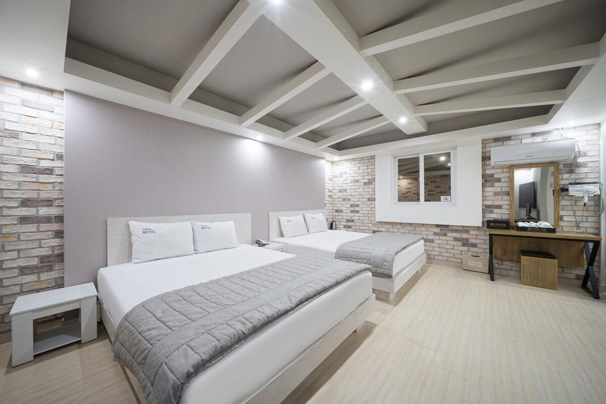 Quarto de hotel com duas camas, uma escrivaninha pequena, um espelho e uma parede de destaque em tijolo.