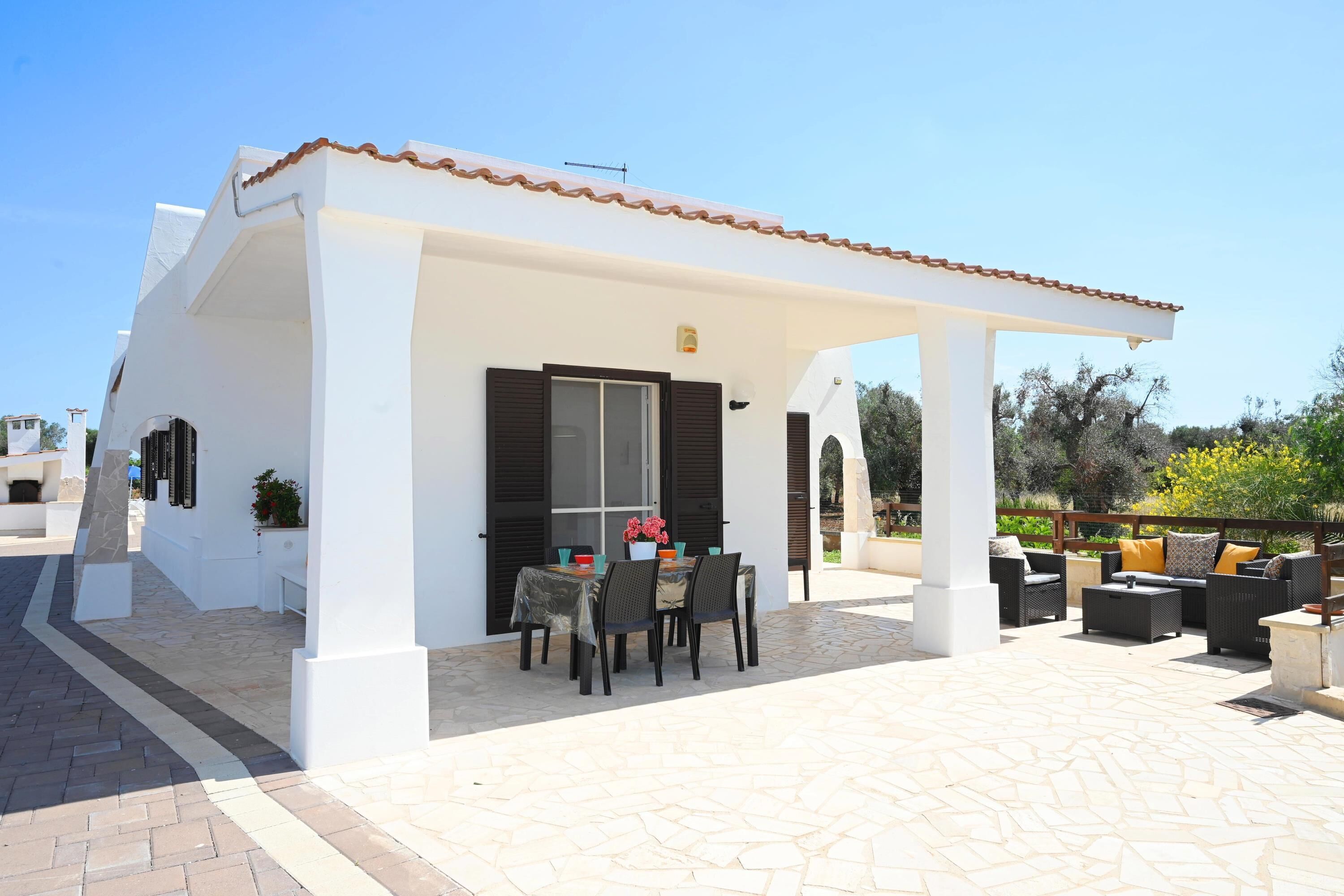 Um pátio externo coberto com mesa e cadeiras, área de estar com sofás, e um edifício branco com telhado de azulejos.