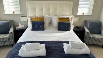 Quarto de hotel com uma cama grande, duas mesas de cabeceira, uma cadeira e uma janela com persianas.
