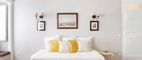 Quarto com cama, roupa de cama branca, duas luminárias fixadas na parede e um quadro emoldurado.