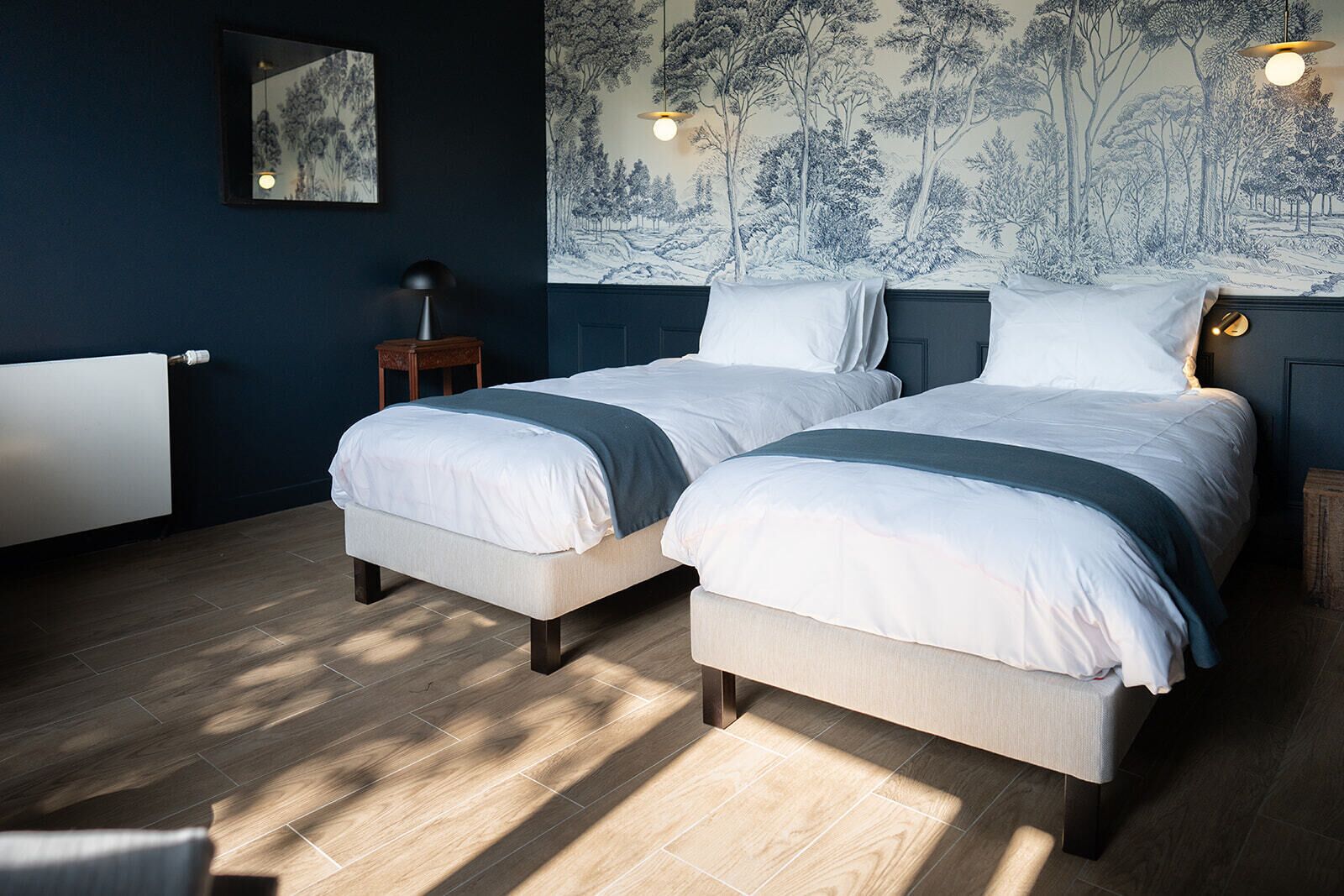 Quarto com duas camas, uma mesinha de cabeceira, um abajur e uma parede com um mural de tema natural.