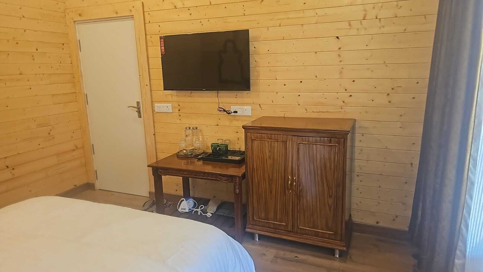 Quarto com uma cama, um armário de madeira, uma mesinha com uma chaleira e xícaras, e uma televisão fixada na parede.