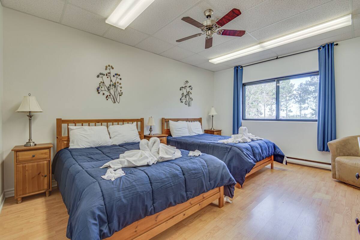 Quarto de hotel com duas camas, cabeceiras de madeira, mesas de cabeceira com luminárias, um ventilador de teto e uma janela com cortinas.