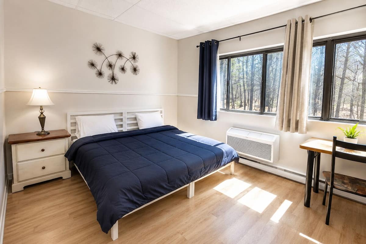 Quarto com cama, mesa de cabeceira, luminária, janela com cortinas, radiador e uma pequena mesa com uma planta.