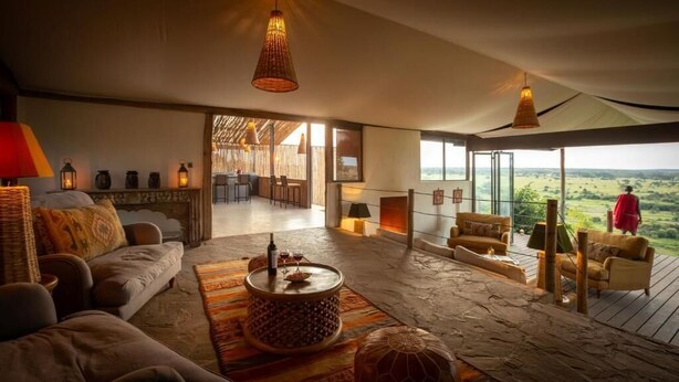 Uma sala de estar espaçosa com uma grande janela que oferece vista para o campo. Há um sofá, uma mesa de centro e uma área de jantar com cadeiras.