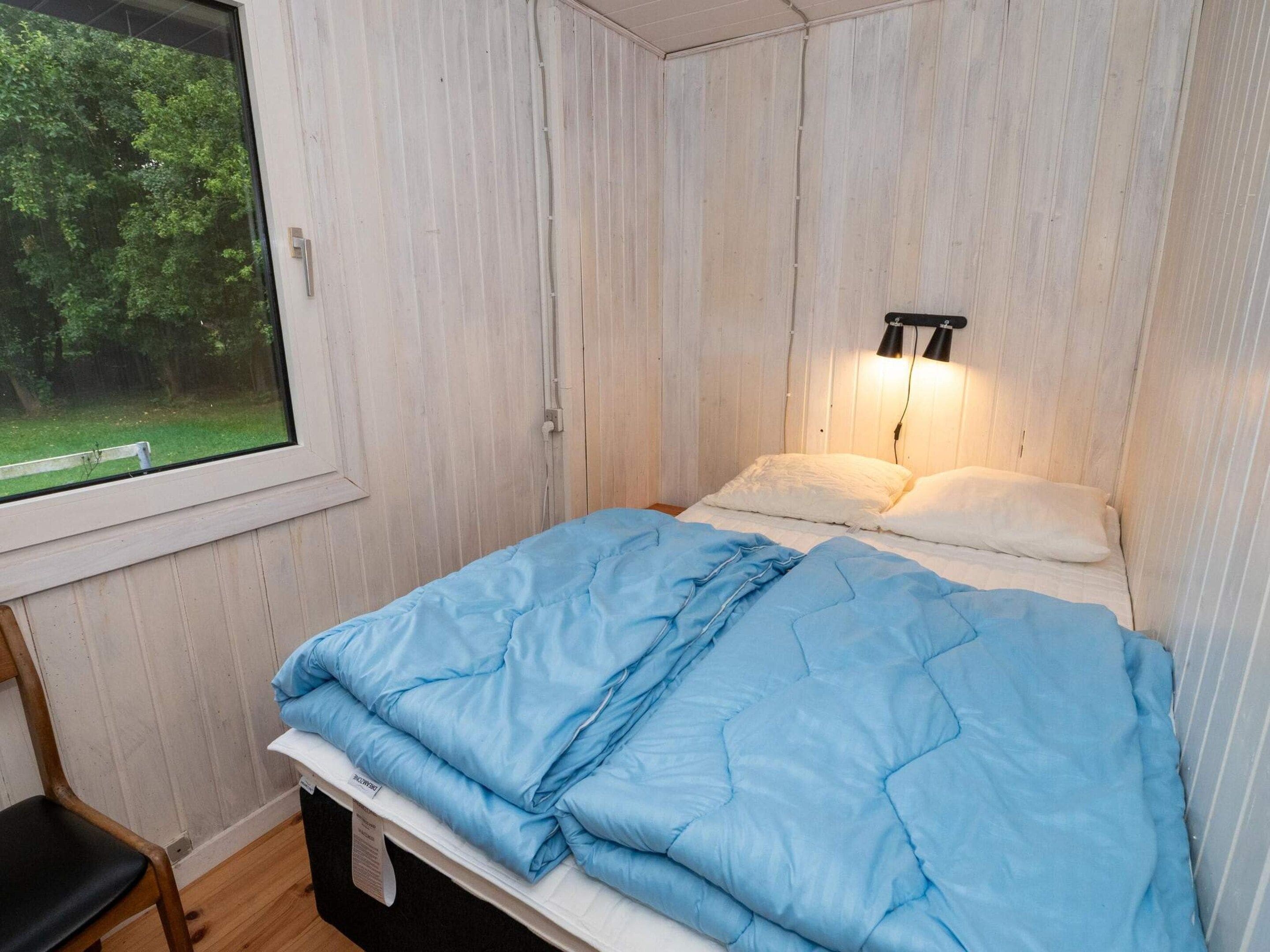 Quarto pequeno com cama, uma cadeira e janela com vista para o verde.