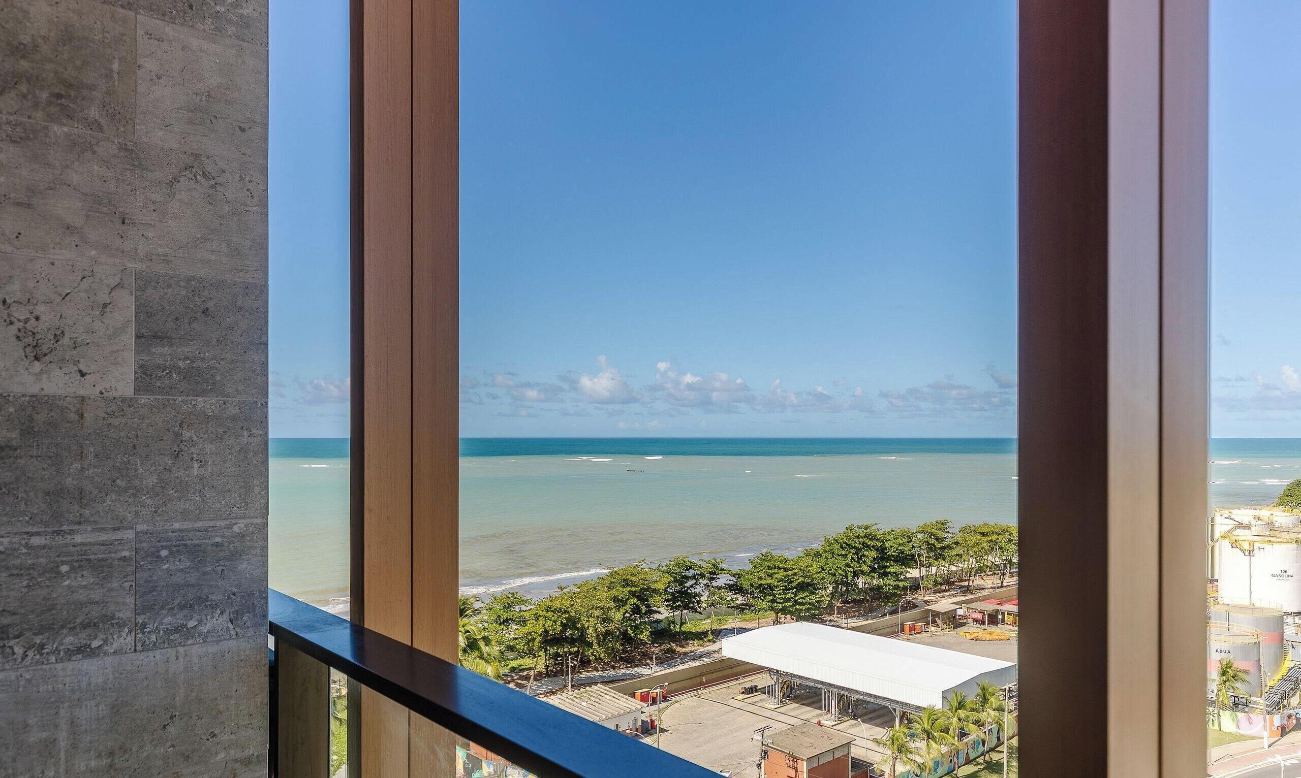 Vista da janela com vista para uma praia sob um céu claro.