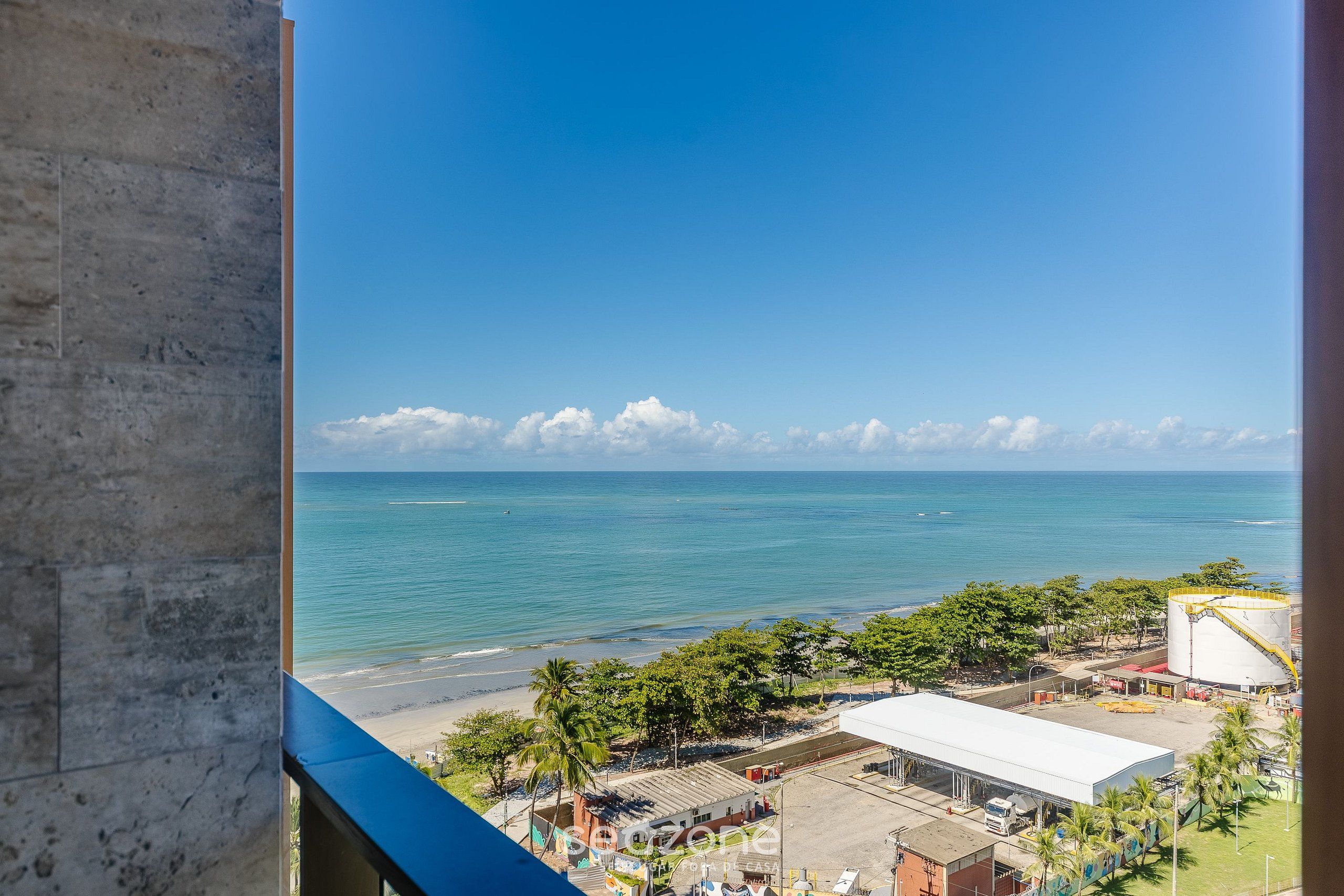 Vista costeira com praia, palmeiras e um edifício branco com telhado plano.