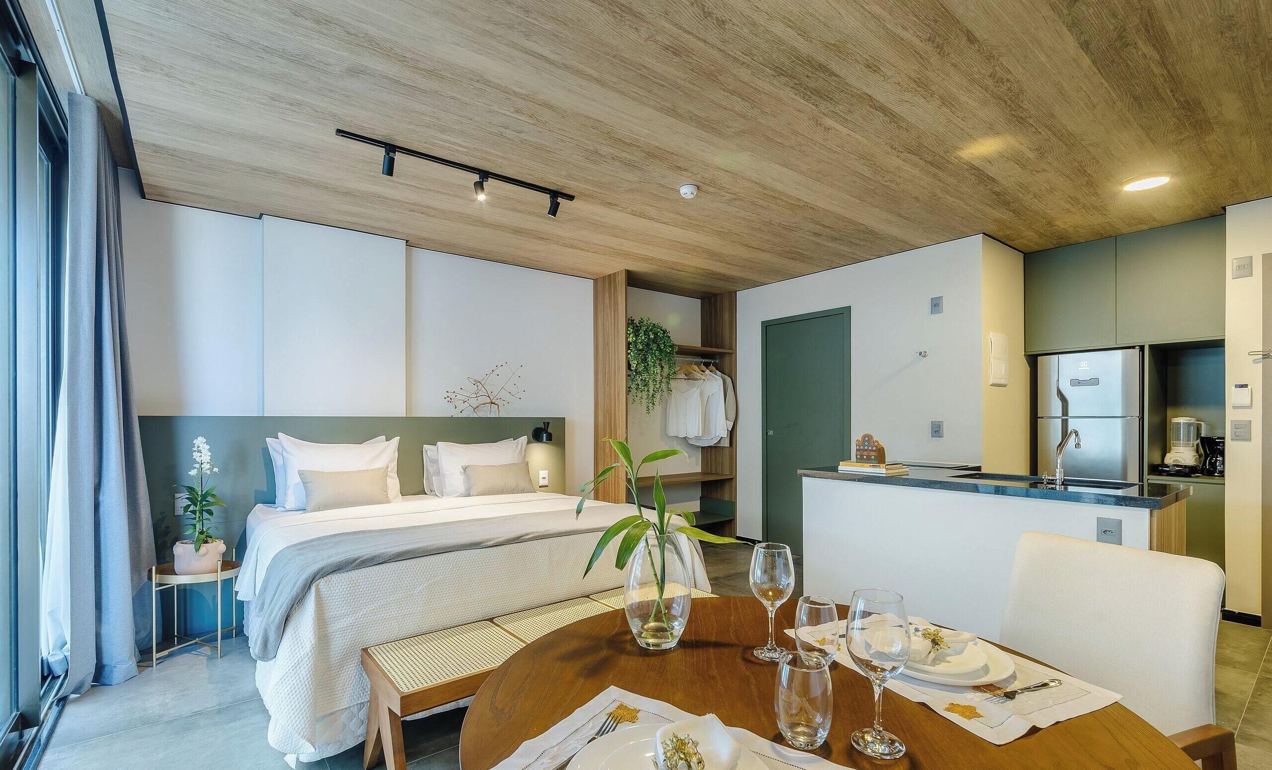 Quarto de hotel com cama, mesa de cabeceira, mesa de jantar para dois, cozinha e banheiro.