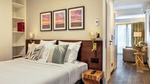 Quarto de hotel com uma cama grande, cabeceira de madeira e vista para o corredor. Há três quadros na parede, uma mesa de cabeceira com abajur e um banquinho de madeira.