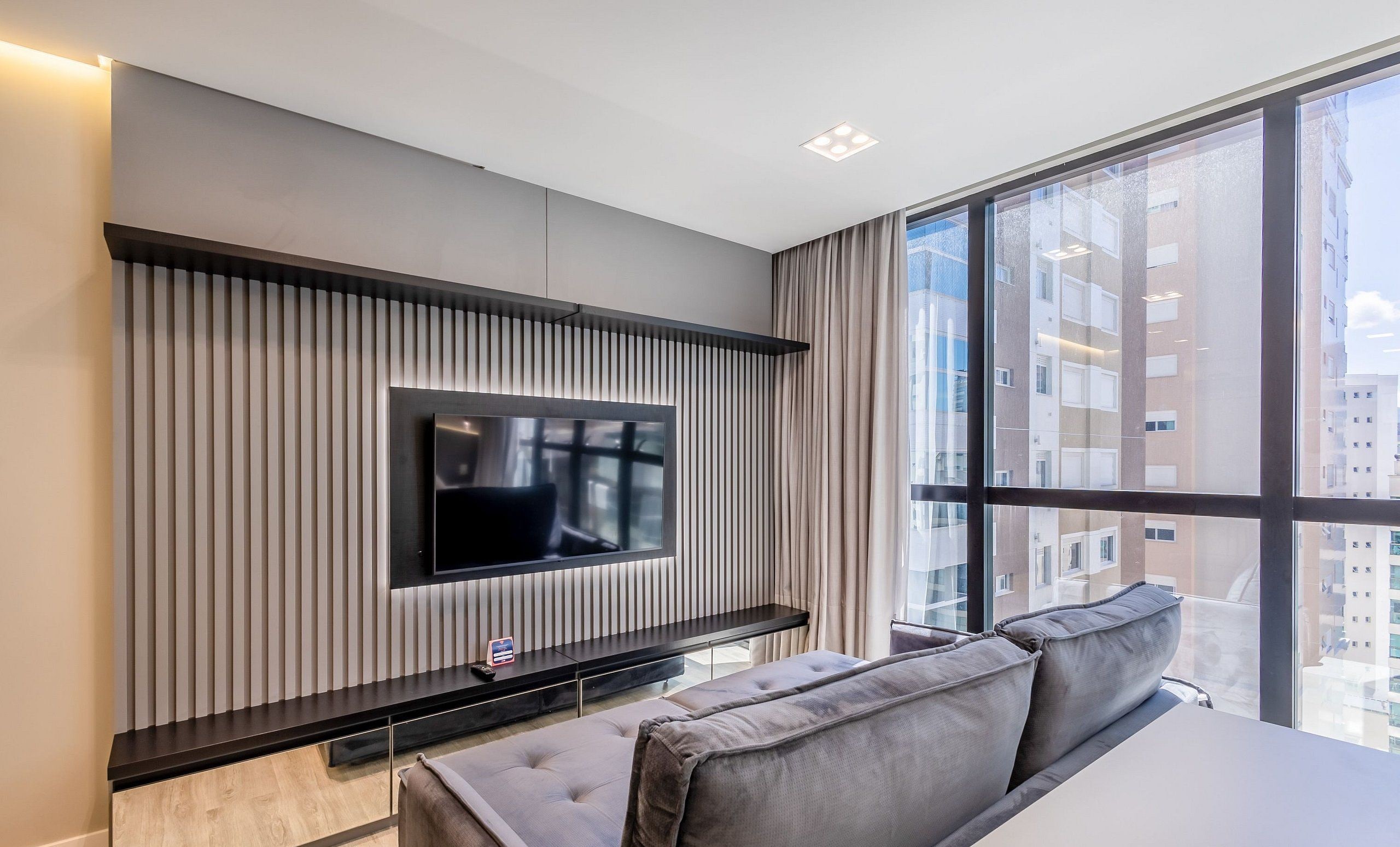 Sala de estar moderna com televisão de tela plana presa na parede, venezianas verticais, sofá e uma janela grande com vista para a cidade.