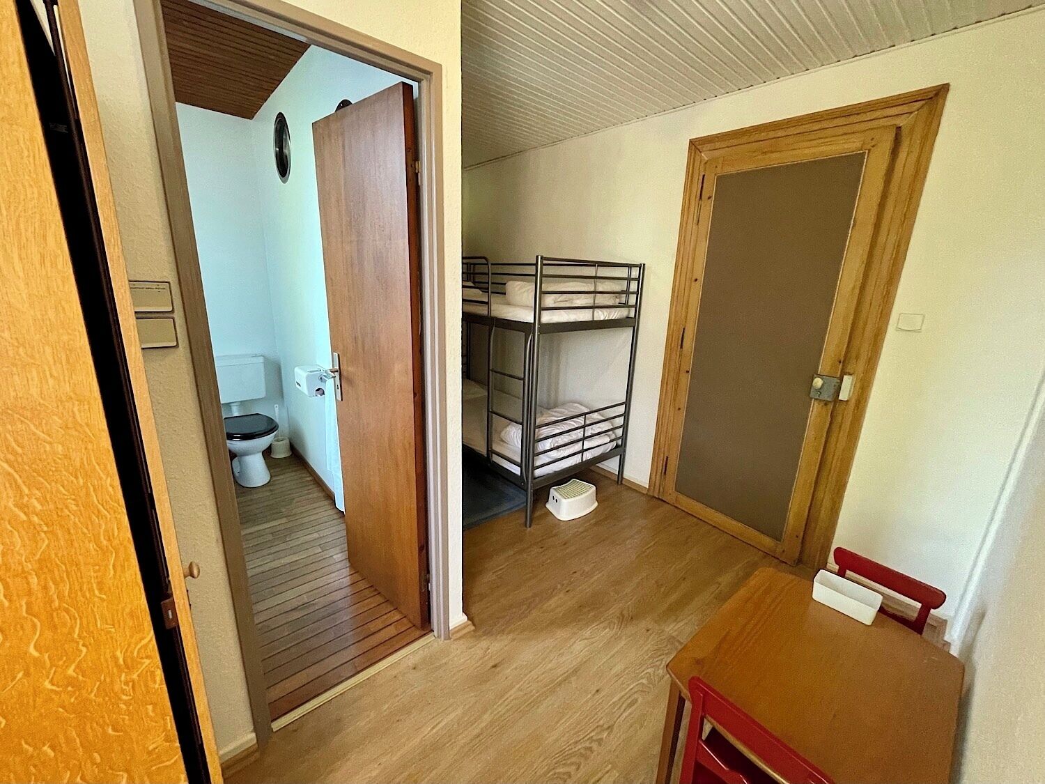 Quarto com piso de madeira, uma cama de solteiro, porta de madeira e uma pequena mesa com uma cadeira.