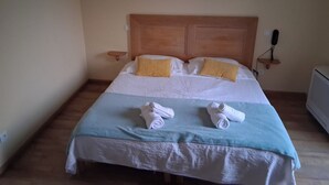 Uma cama de casal com cabeceira de madeira, duas mesas de cabeceira com abajures e um pequeno prateleira na parede.