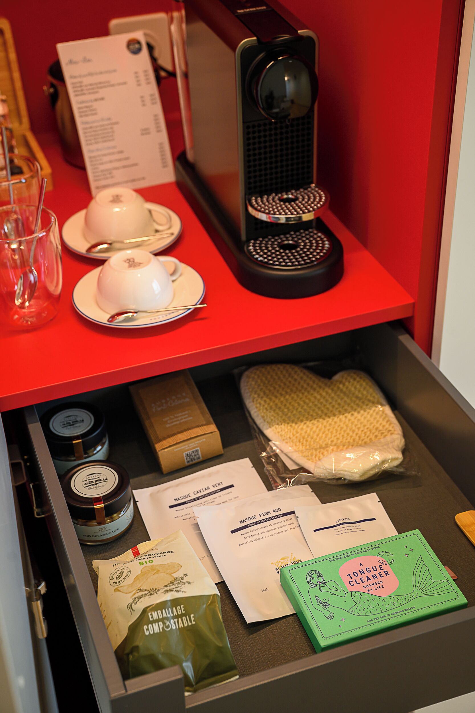 Minibar de quarto de hotel com bancada vermelha, cafeteira, chaleira e diversos itens como uma caixa verde, um saco de sacos de lixo compostáveis e alguns papéis.