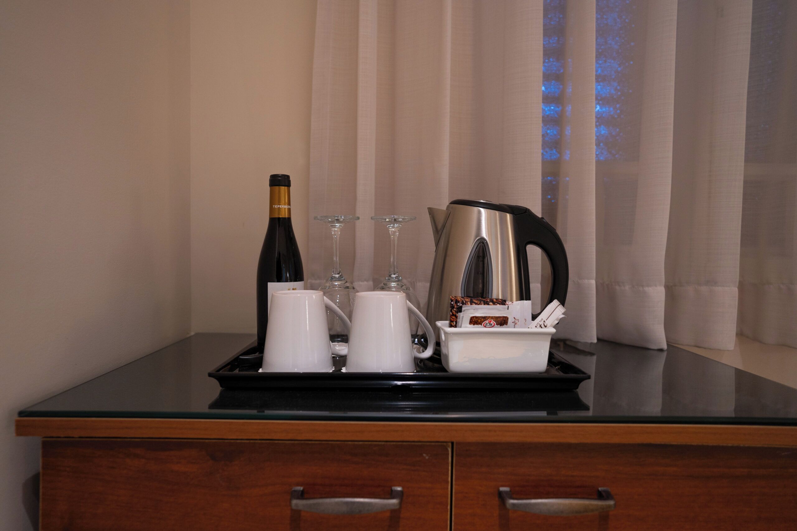 Quarto de hotel com um armário de madeira, uma bandeja com uma garrafa de vinho, duas taças de vinho, uma cafeteira e uma seleção de petiscos.