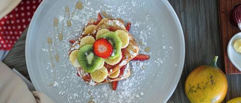 Um prato de panquecas coberto com frutas e açúcar em pó, um copo de sumo de laranja e uma chávena de café sobre uma mesa de madeira.
