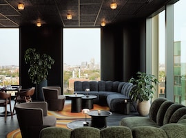 Espaço lounge moderno com sofá, poltronas e mesa de centro, com vista para a cidade através de grandes janelas.