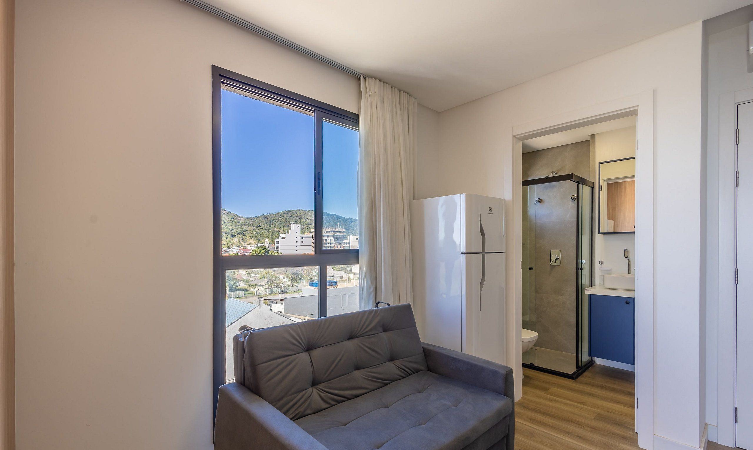 Um apartamento moderno com sofá, geladeira, box de vidro e vista para prédios e colinas.