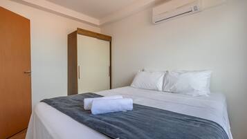 Quarto de hotel com uma cama, roupa de cama branca, um cobertor cinza, uma porta de madeira e um ar condicionado.
