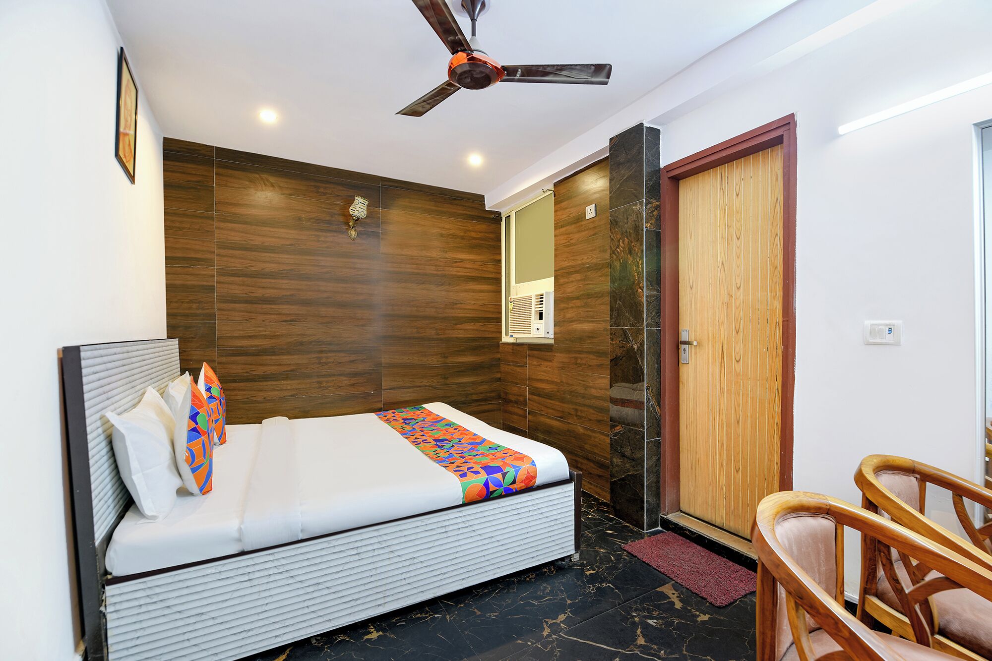 Quarto com cama, ventoinha no teto, porta de madeira e uma cadeira.