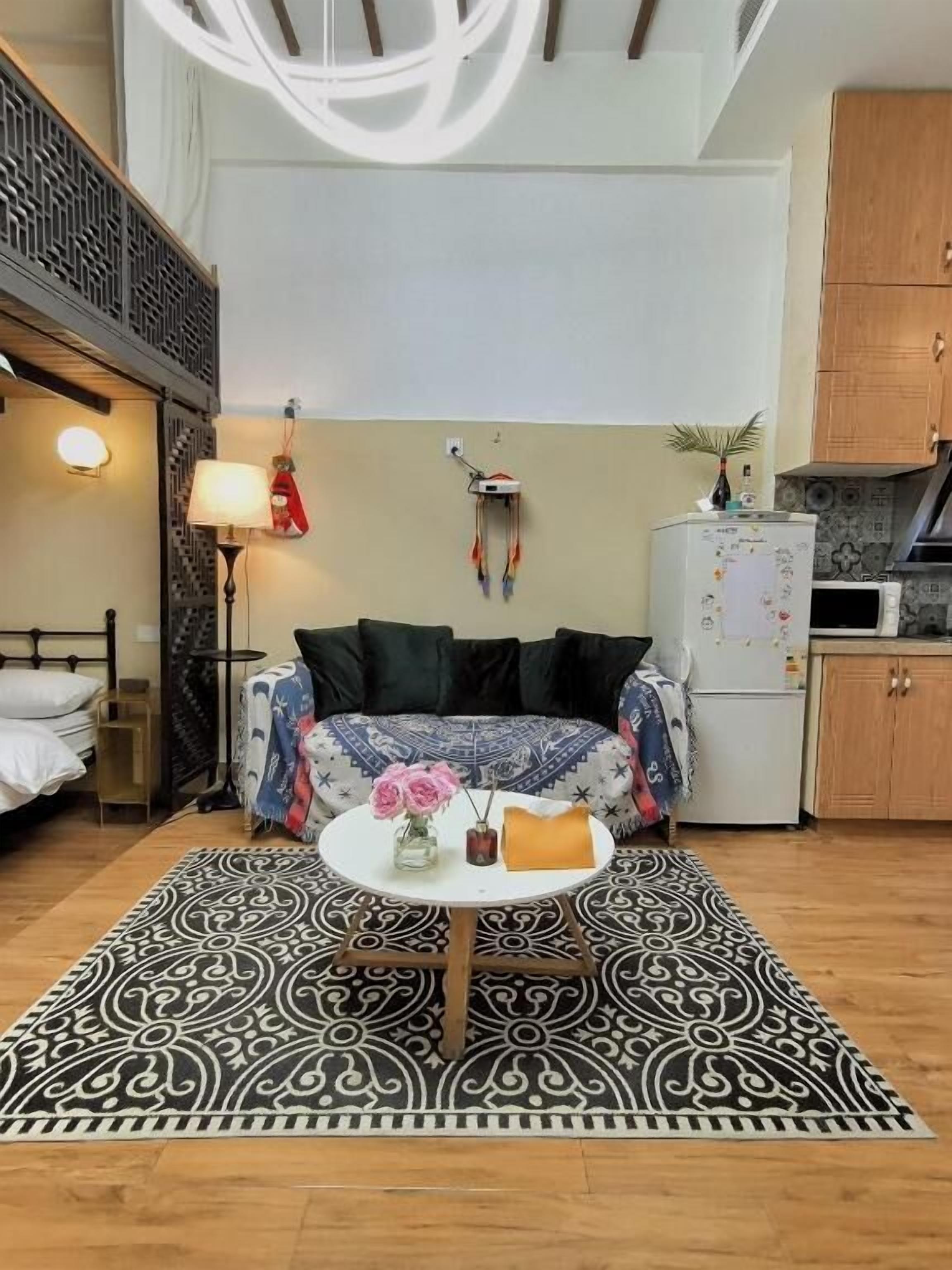 Um espaço compacto com cama, sofá, mesa redonda pequena com um vaso de flores e área de cozinha com armários e geladeira.