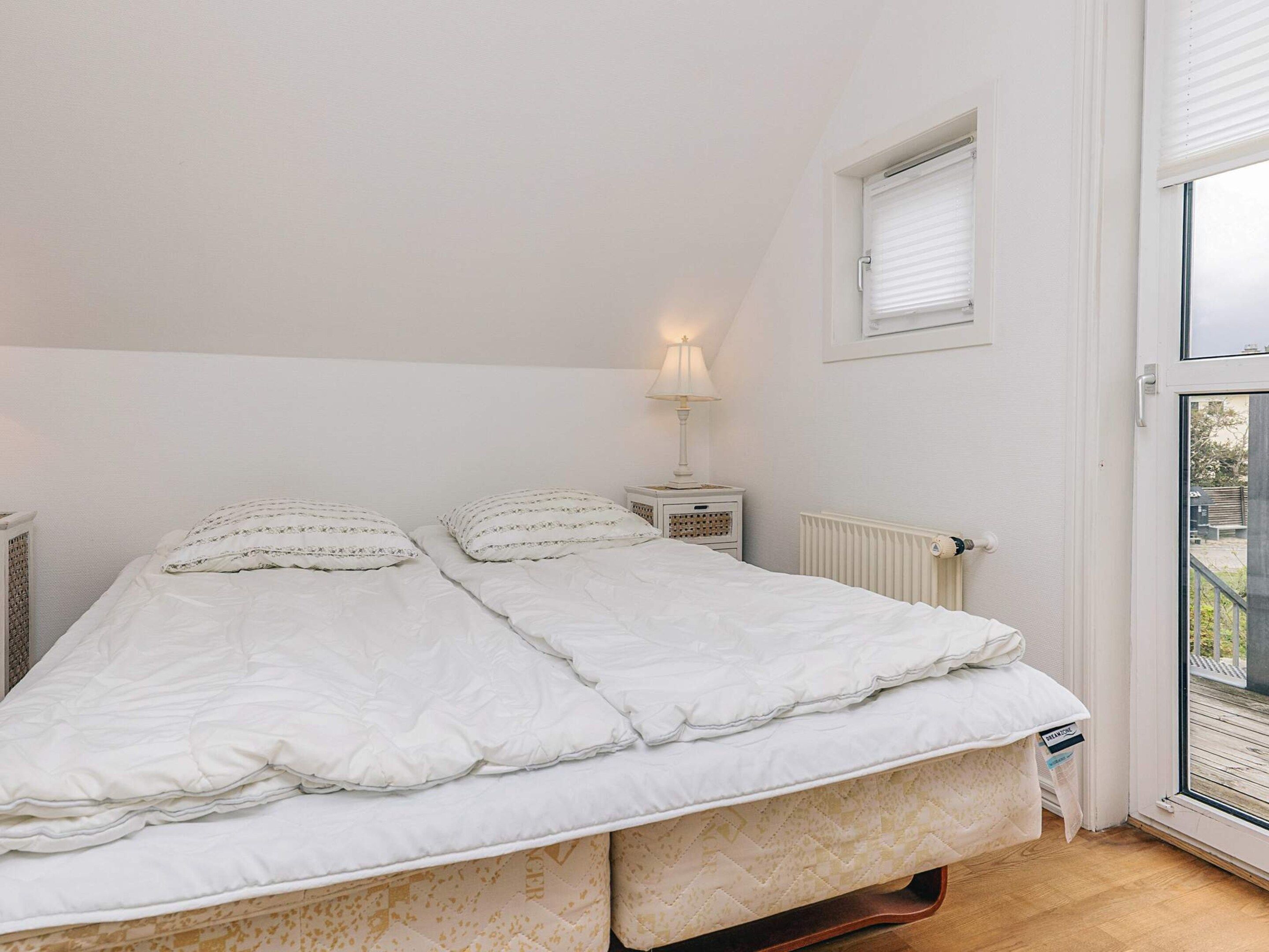 Quarto com cama, mesa de cabeceira com abajur, janela com persianas e radiador.